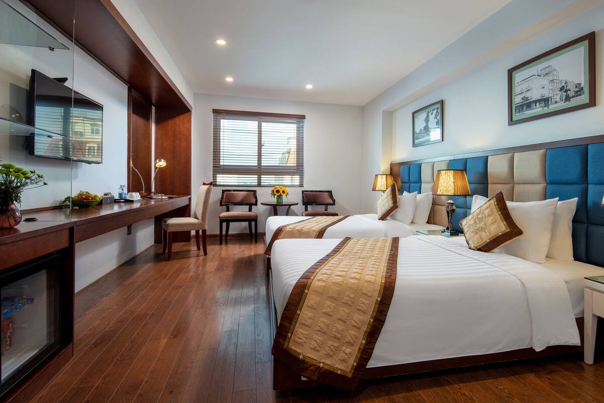 Quarto de hotel com duas camas, uma escrivaninha, cadeiras e uma televisão.