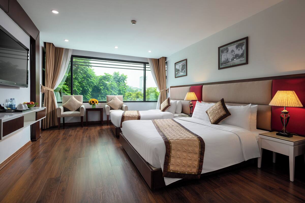 Quarto de hotel com duas camas, televisão, escrivaninha e janela com vista para árvores.