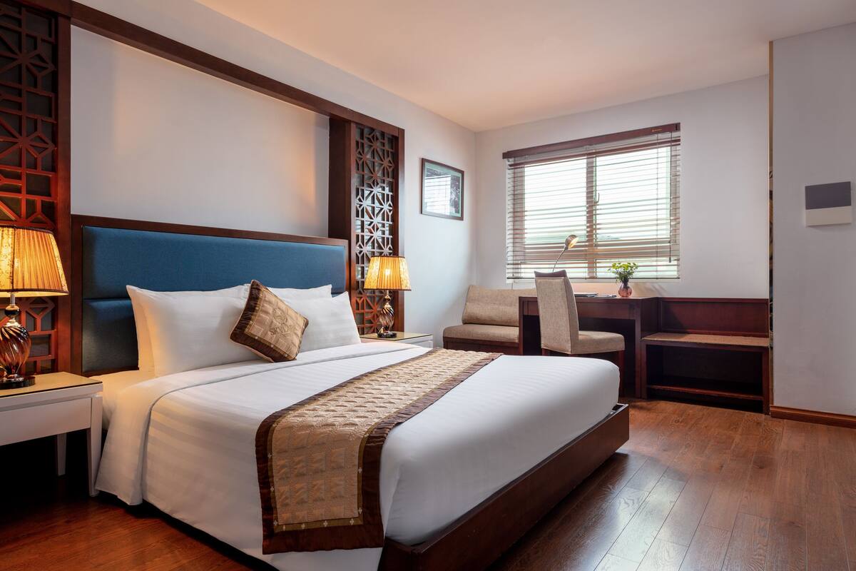 Quarto de hotel com uma cama grande, uma escrivaninha, uma cadeira e uma janela com persianas.