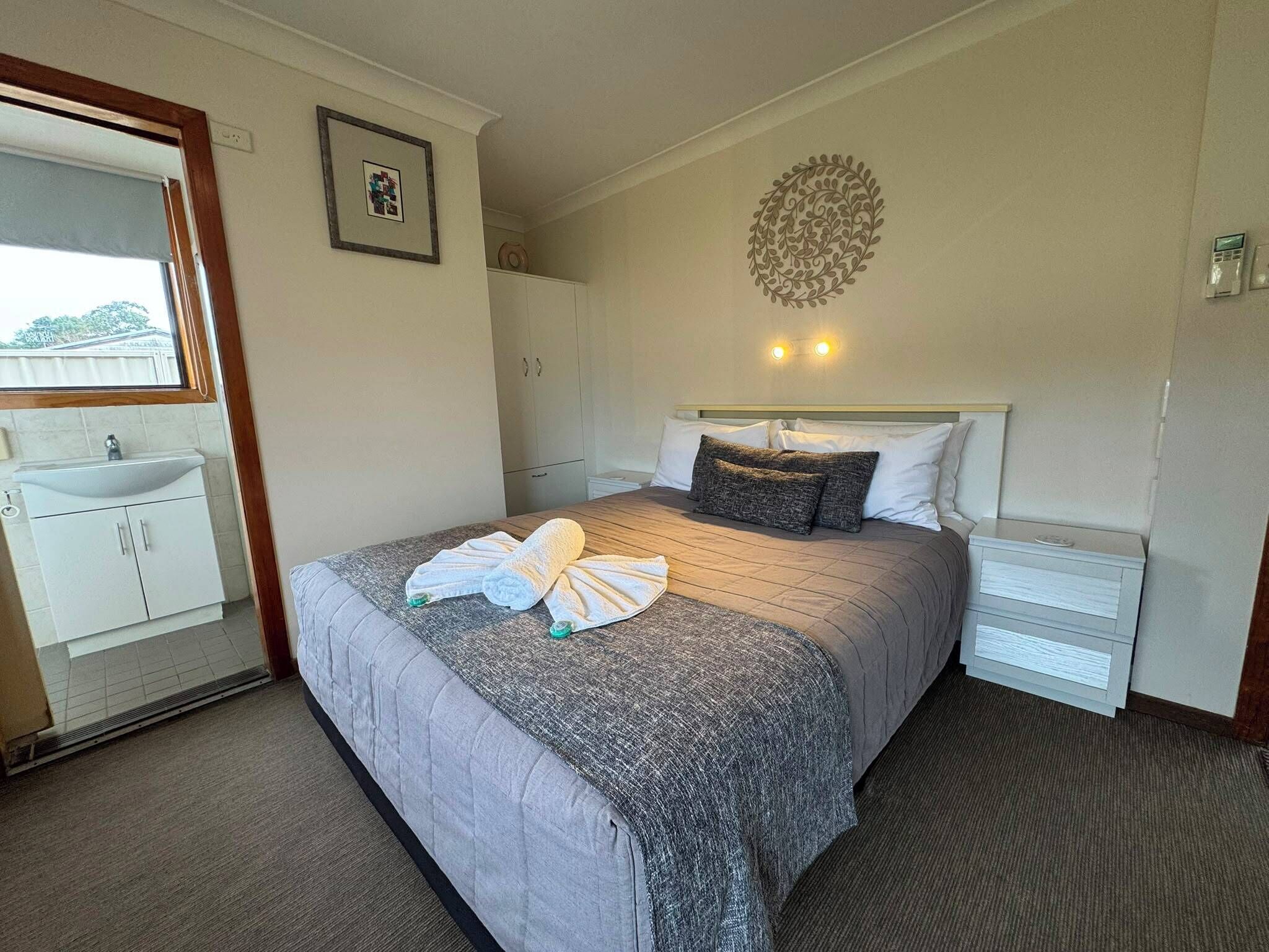 Quarto com cama, mesa de cabeceira, luminária na parede, decoração na parede, janela com vista e banheiro com pia e armário.