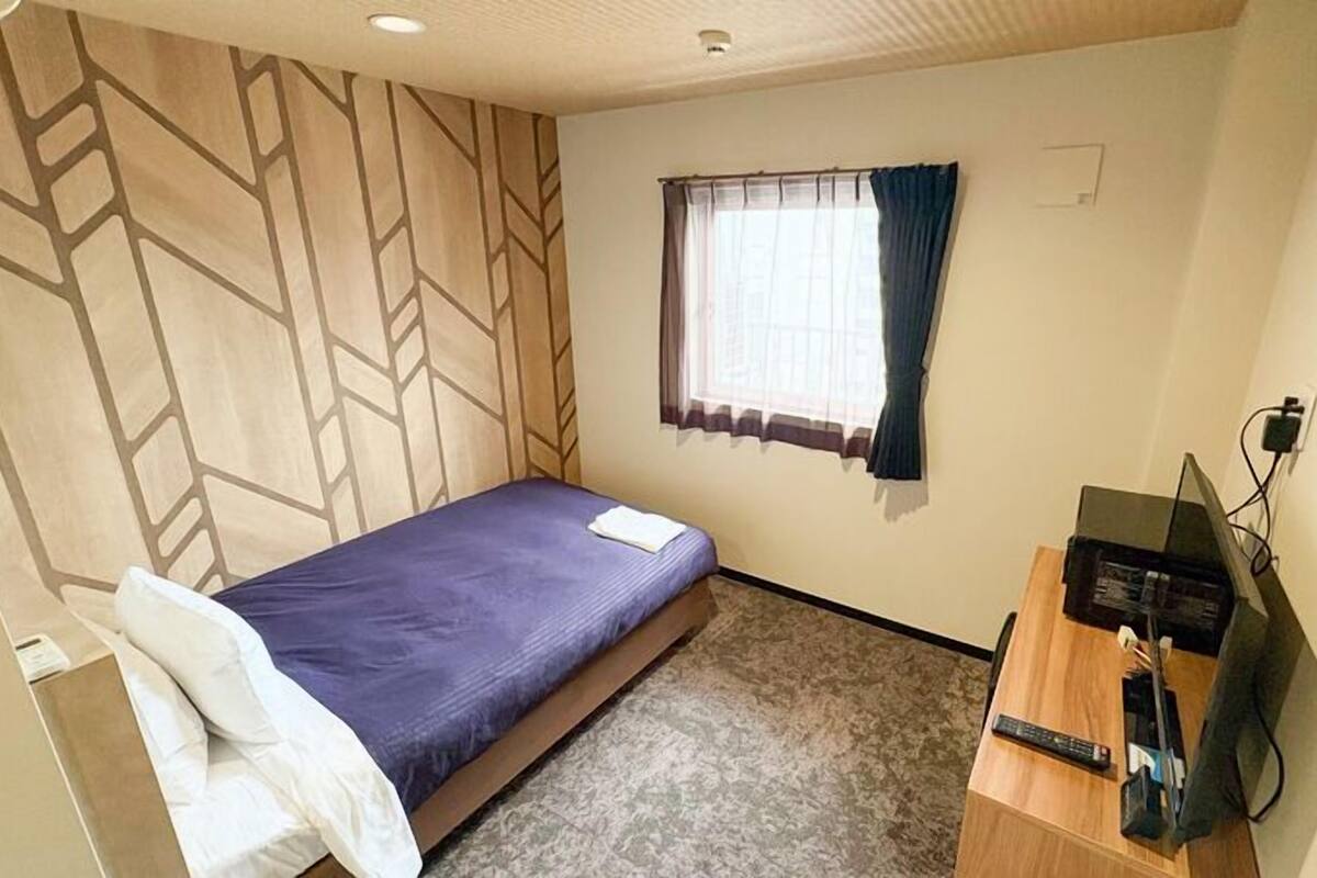 Quarto pequeno com uma cama de solteiro, cabeceira de madeira, janela com cortinas, escrivaninha com computador e televisão.