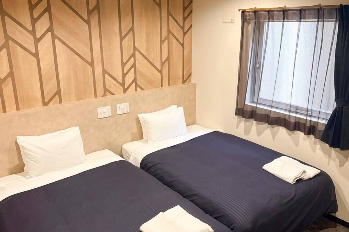 Quarto de hotel com duas camas, uma janela com cortinas e um design geométrico na parede.