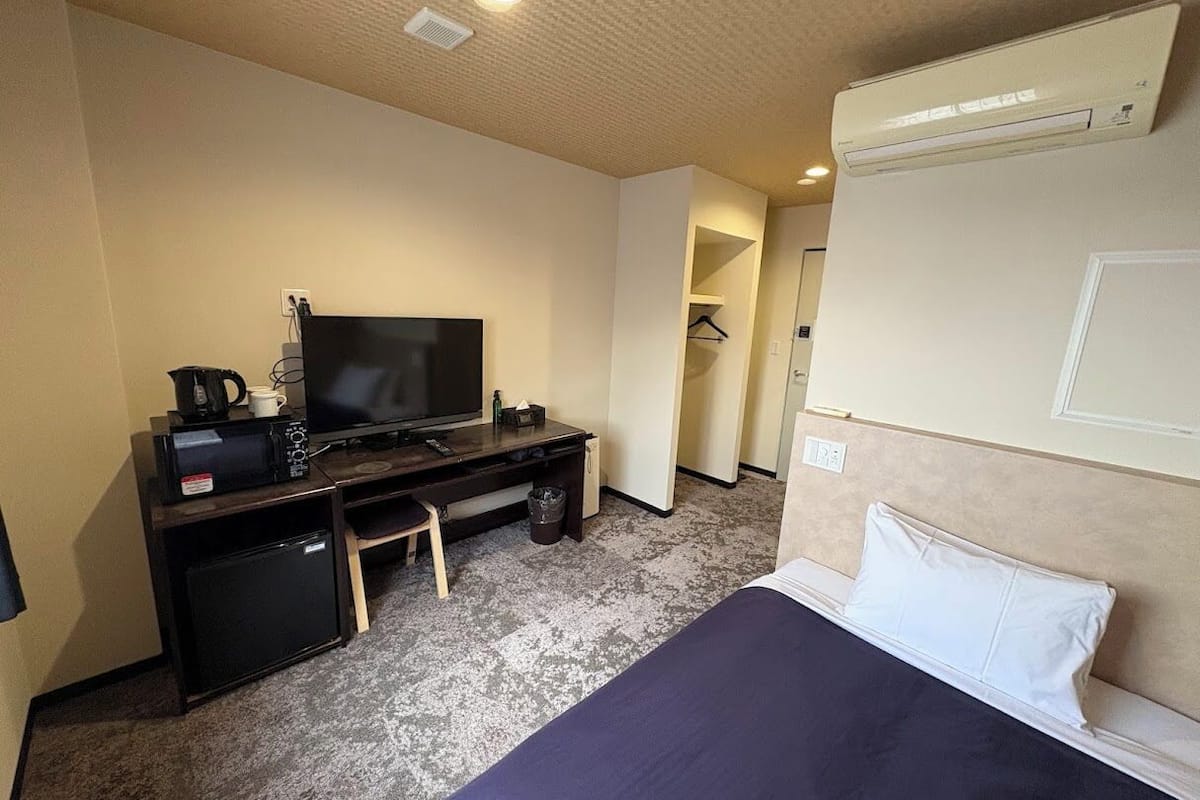 Quarto de hotel com cama, escrivaninha com televisão, micro-ondas e pequena geladeira.