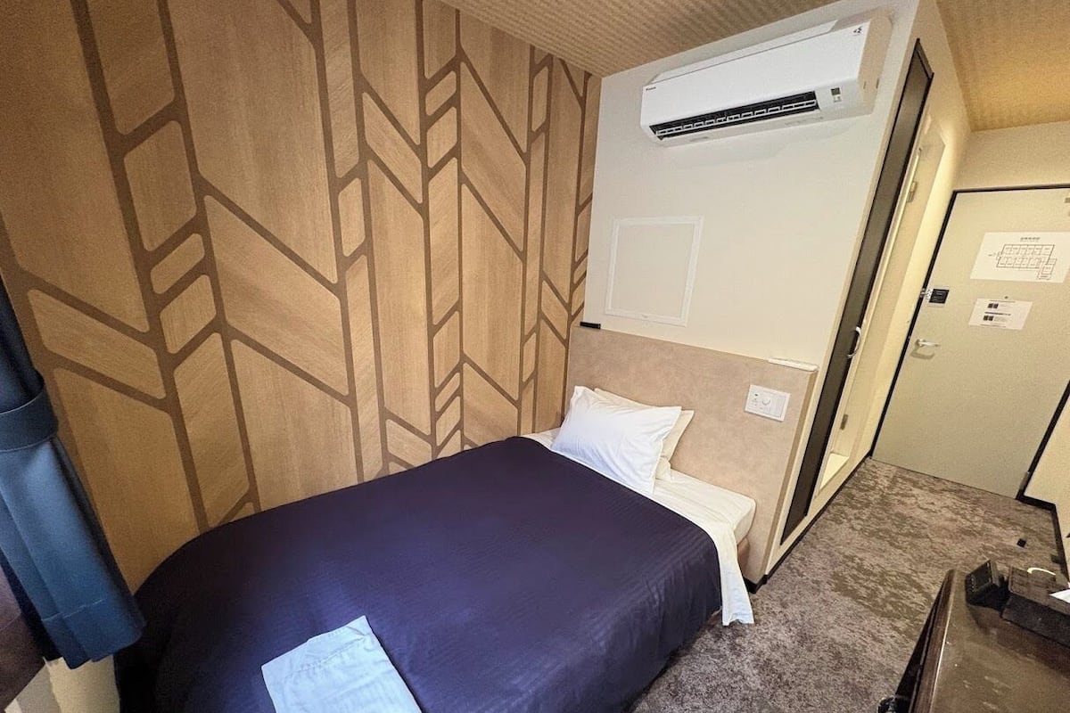 Quarto de hotel com cama, ar condicionado fixado na parede, porta com fechadura e pequena mesa com cadeira.