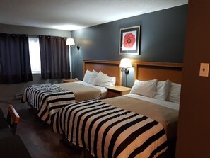 Quarto de hotel com duas camas, uma mesa de cabeceira com abajur, um banco e uma grande pintura de flores na parede.