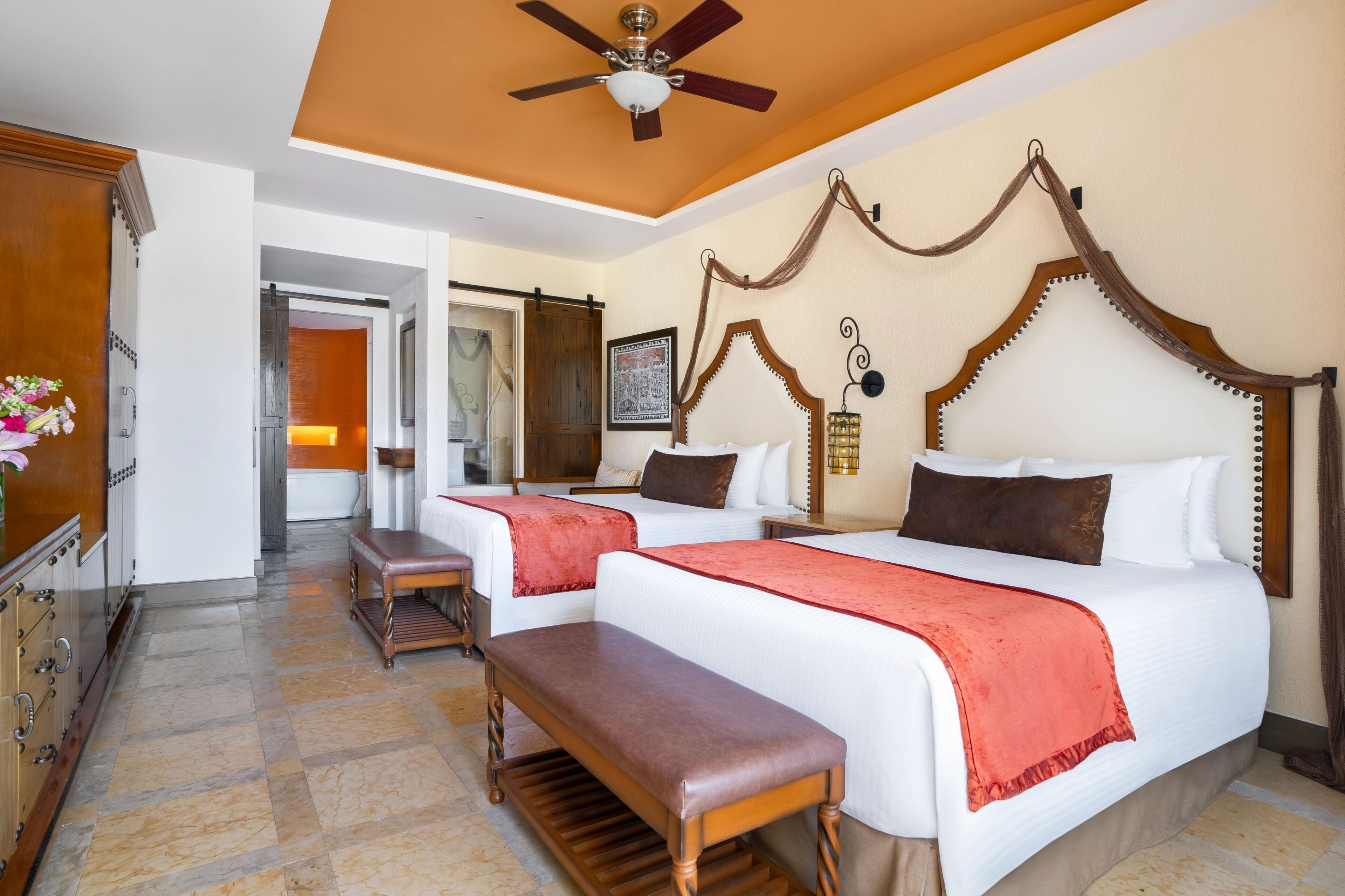 Quarto de hotel com duas camas, um banco de madeira, um ventilador de teto e um guarda-roupa de madeira.