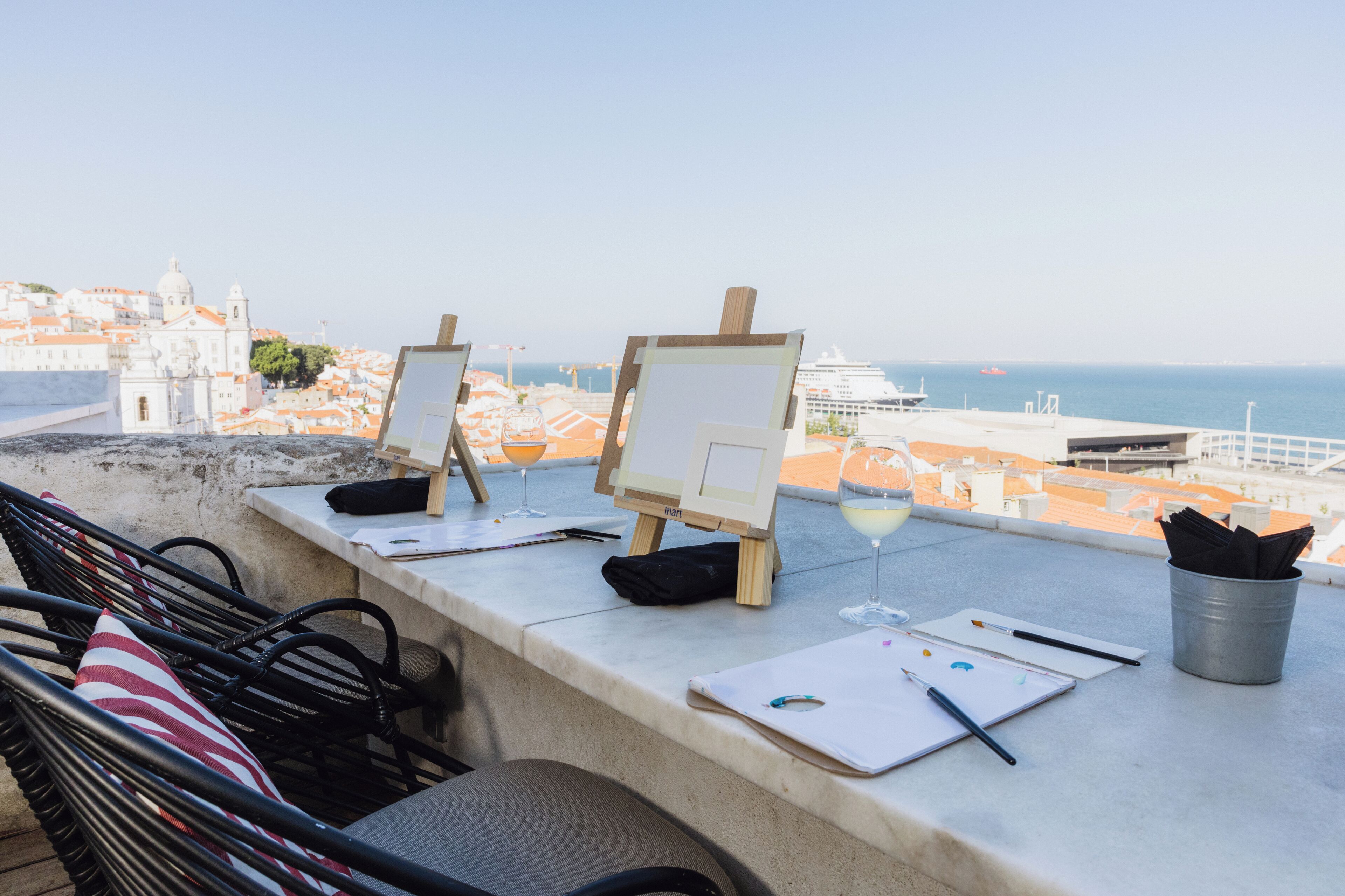 Terraço com cavaletes e materiais de arte, com vista para uma paisagem costeira com edifícios históricos e um navio de cruzeiro.