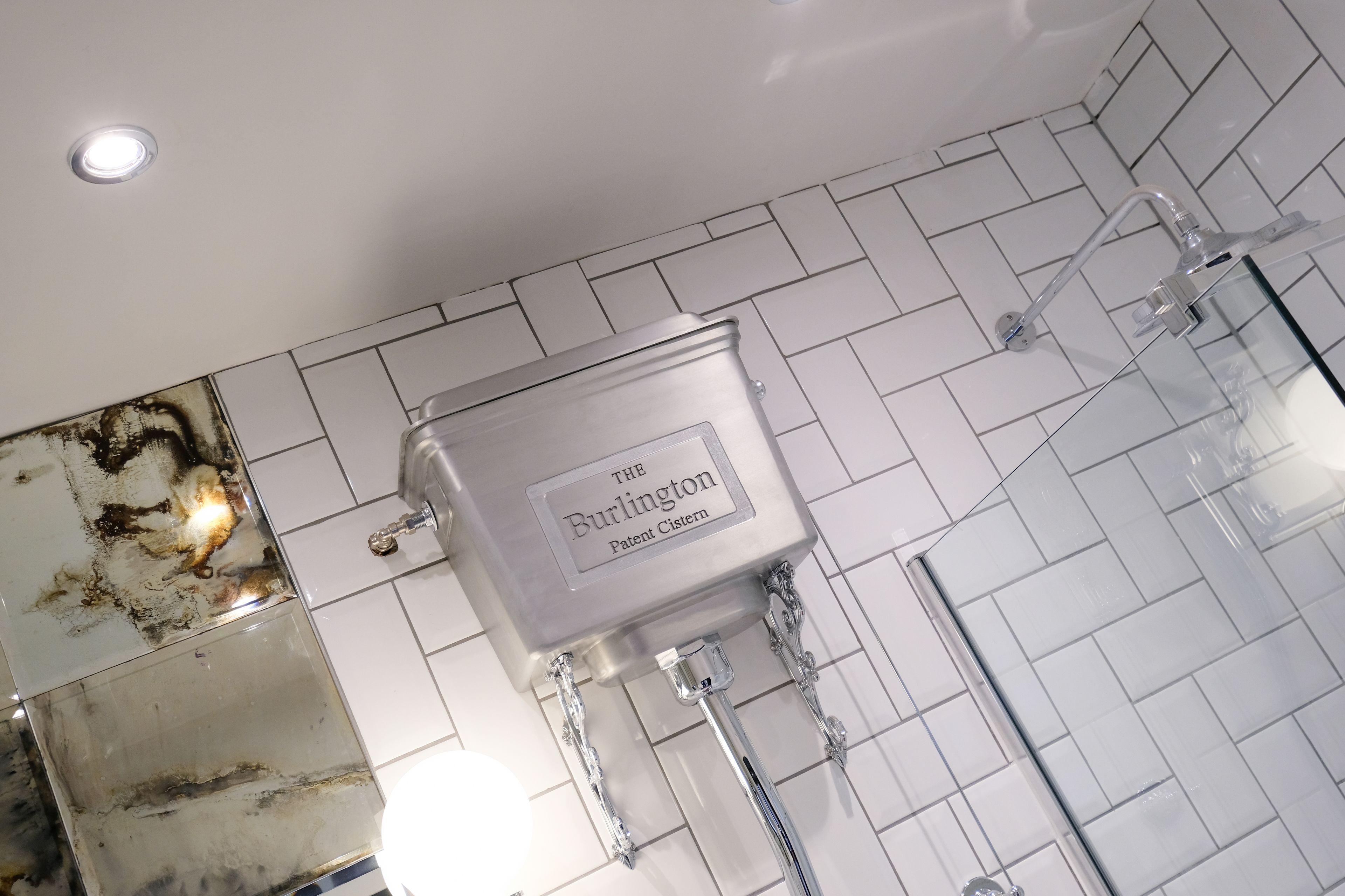 Casa de banho com paredes revestidas de azulejos brancos, um espelho danificado e um termoacumulador com a inscrição "The Burlington Patent Cistern".