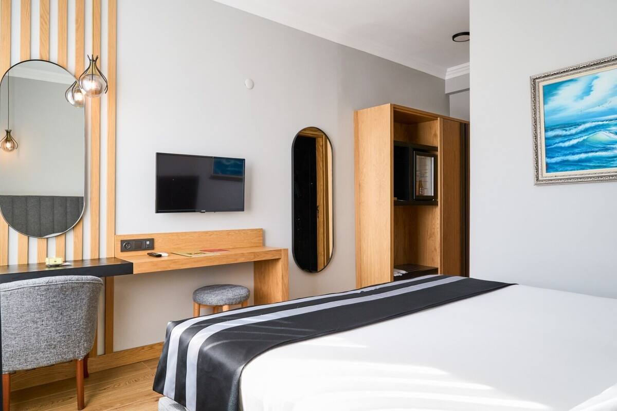 Quarto de hotel moderno com cama, escrivaninha, cadeira, televisão, espelho grande e guarda-roupa.