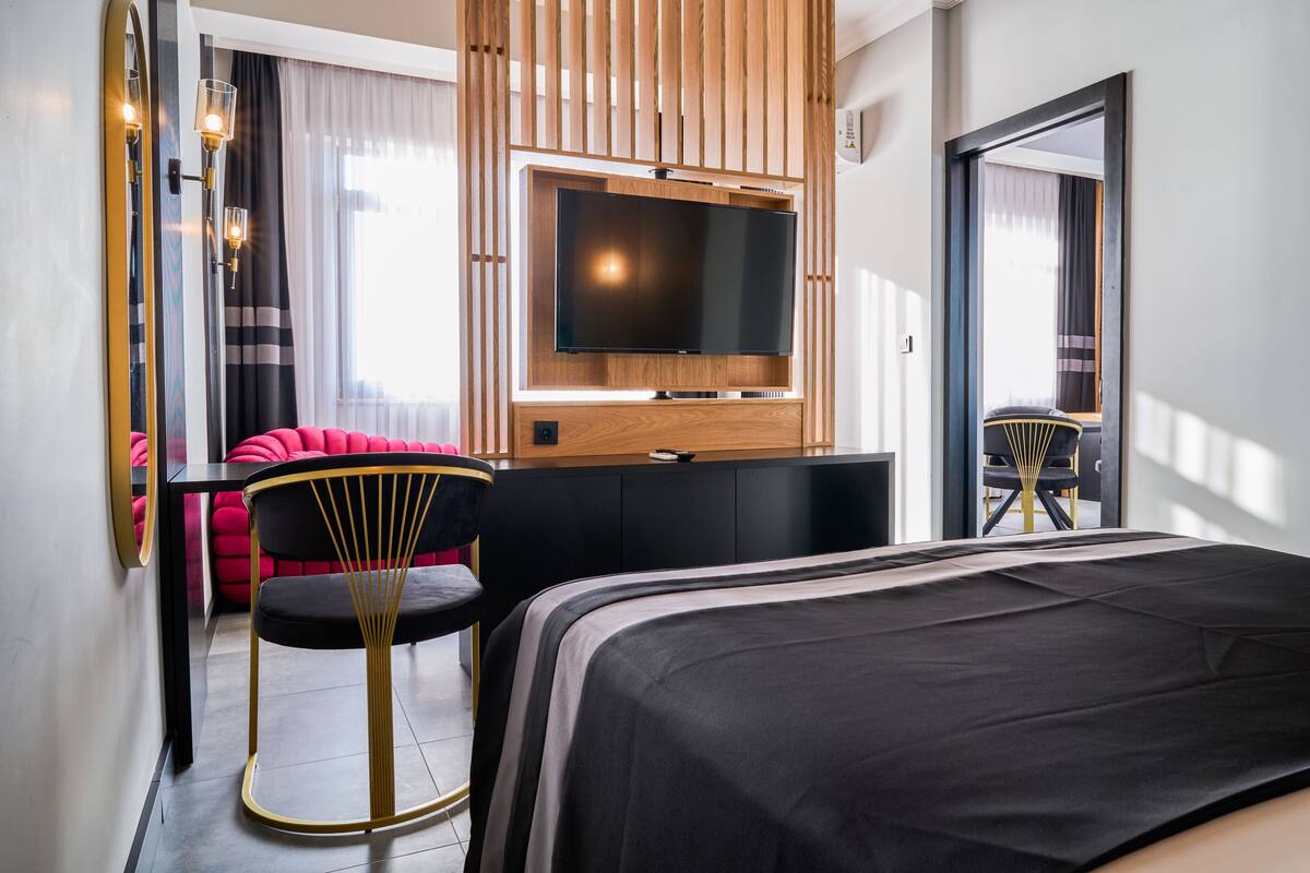 Quarto de hotel moderno com uma cama grande, uma TV fixada em um painel de madeira, uma escrivaninha com cadeira e um espelho amplo.