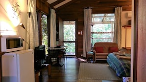 Quarto compacto com teto de madeira, mesa de jantar com cadeiras, sofá, cama com manta xadrez e micro-ondas sobre uma geladeira pequena.