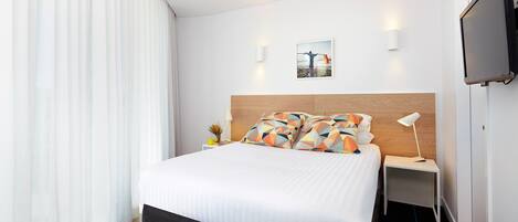 Quarto de hotel moderno com uma cama grande, uma televisão de ecrã plano na parede e uma pequena mesa de cabeceira com uma luminária.