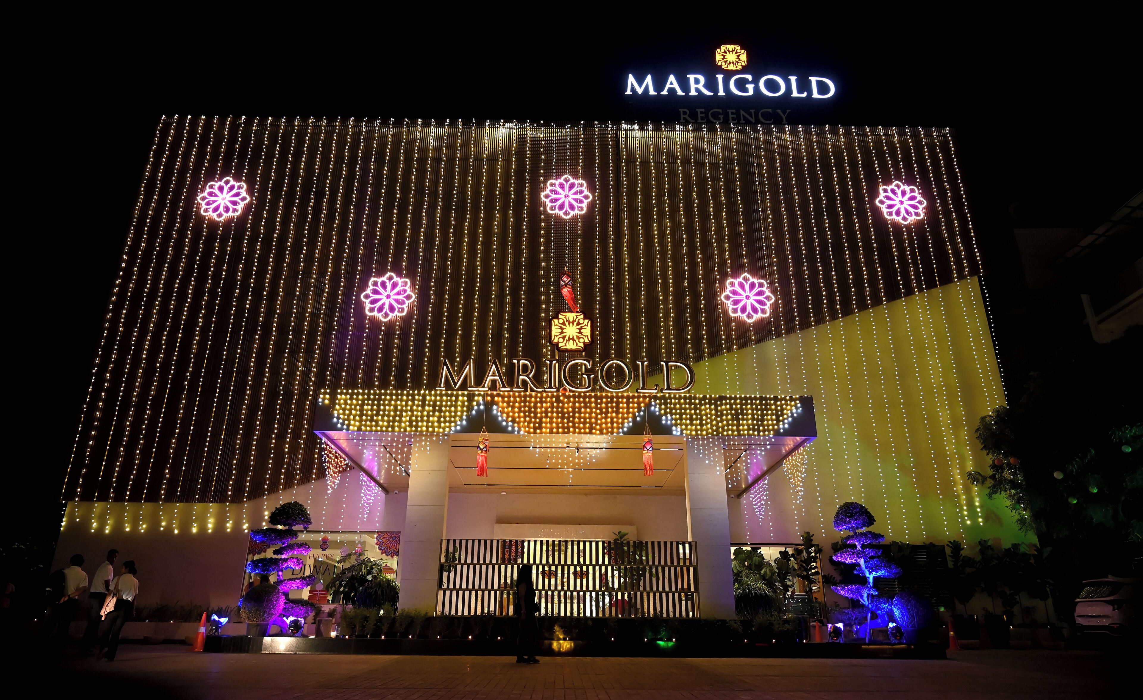 Fachada de um hotel Marigold à noite, com luzes de Natal iluminadas e detalhes florais.