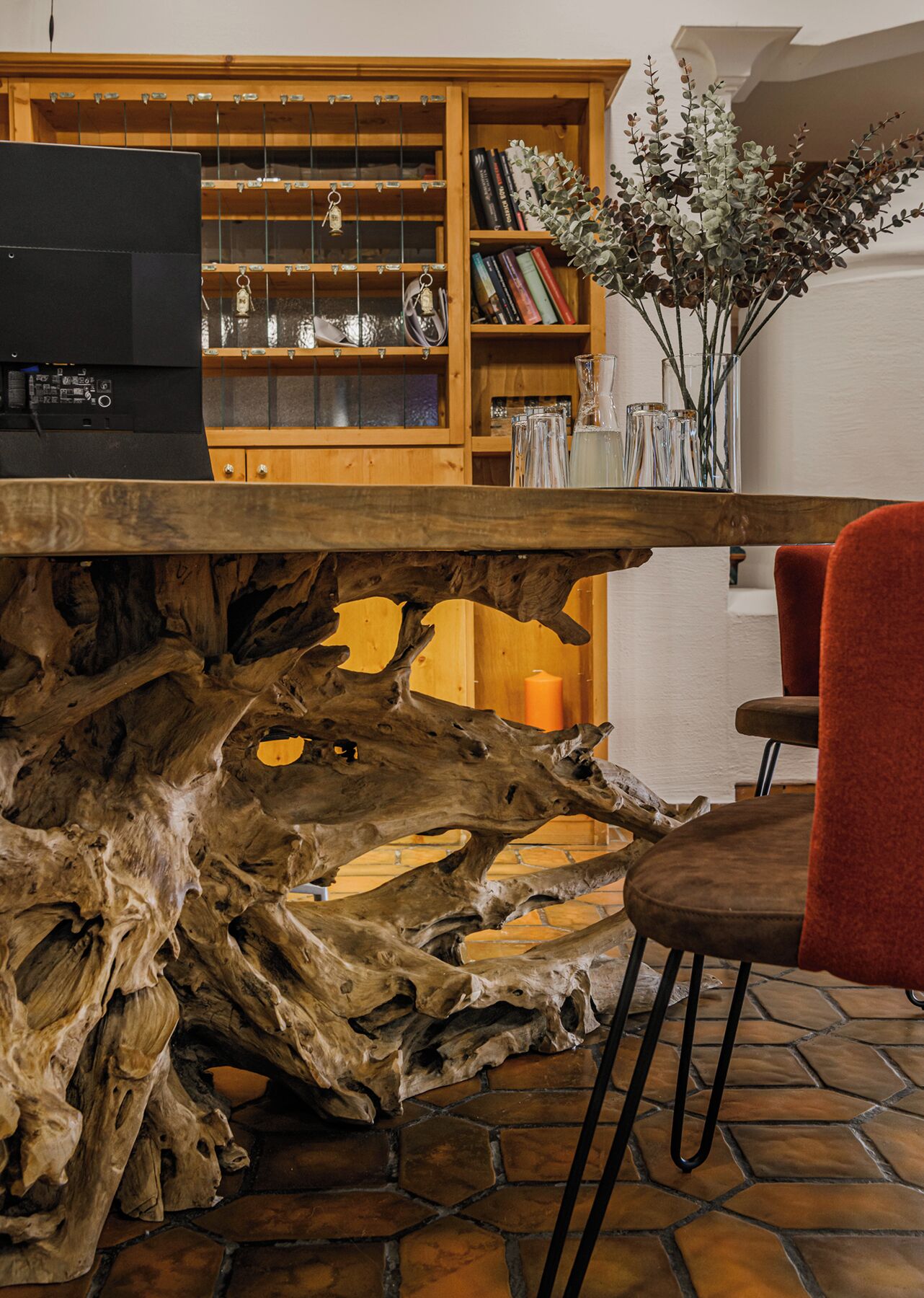 Uma mesa de madeira rústica com design singular, uma cadeira vermelha, uma estante com livros e objetos decorativos, e um vaso com ramos.