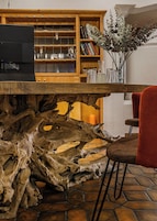 Uma mesa de madeira rústica com design singular, uma cadeira vermelha, uma estante com livros e objetos decorativos, e um vaso com ramos.