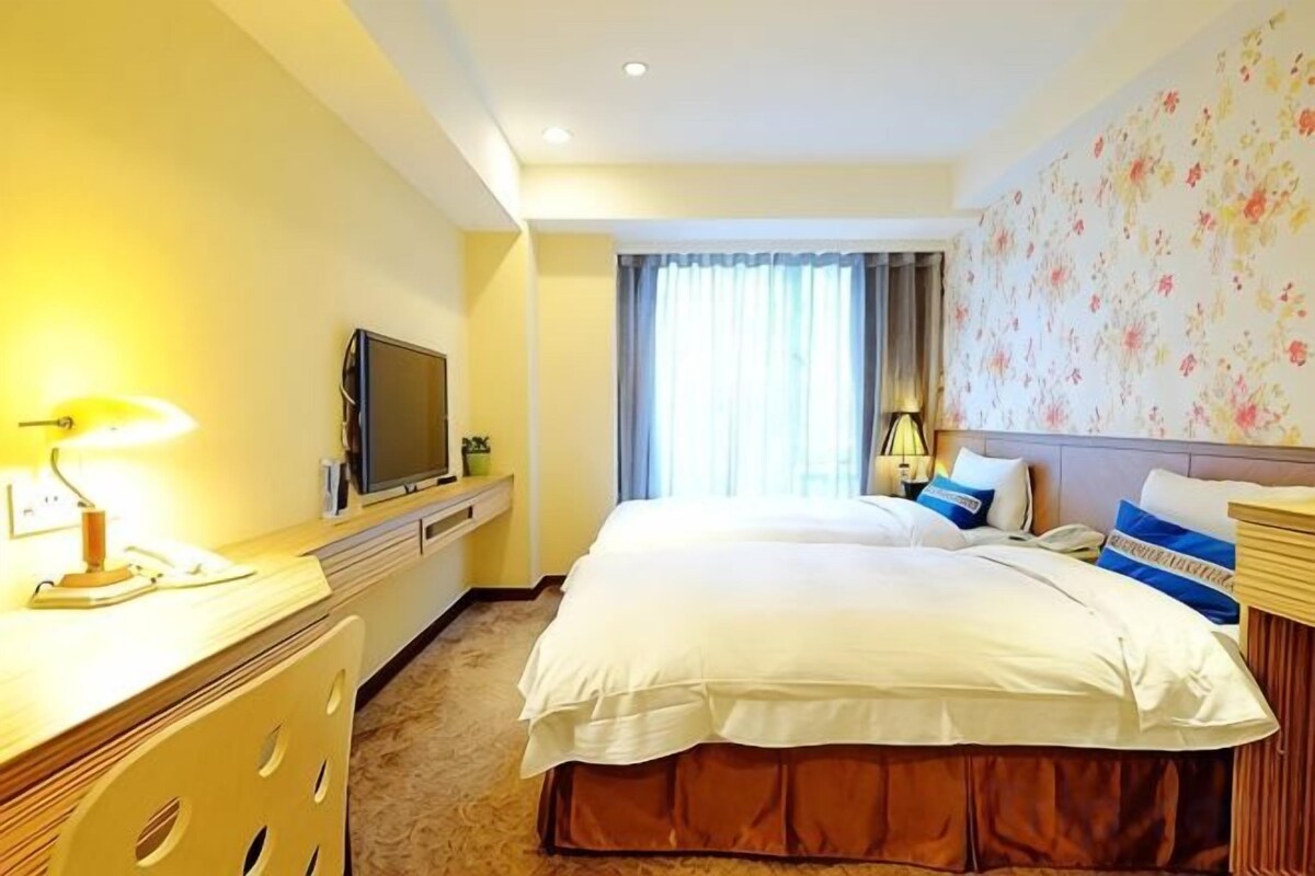 Quarto de hotel com cama, escrivaninha com luminária, televisão e janela com cortinas.