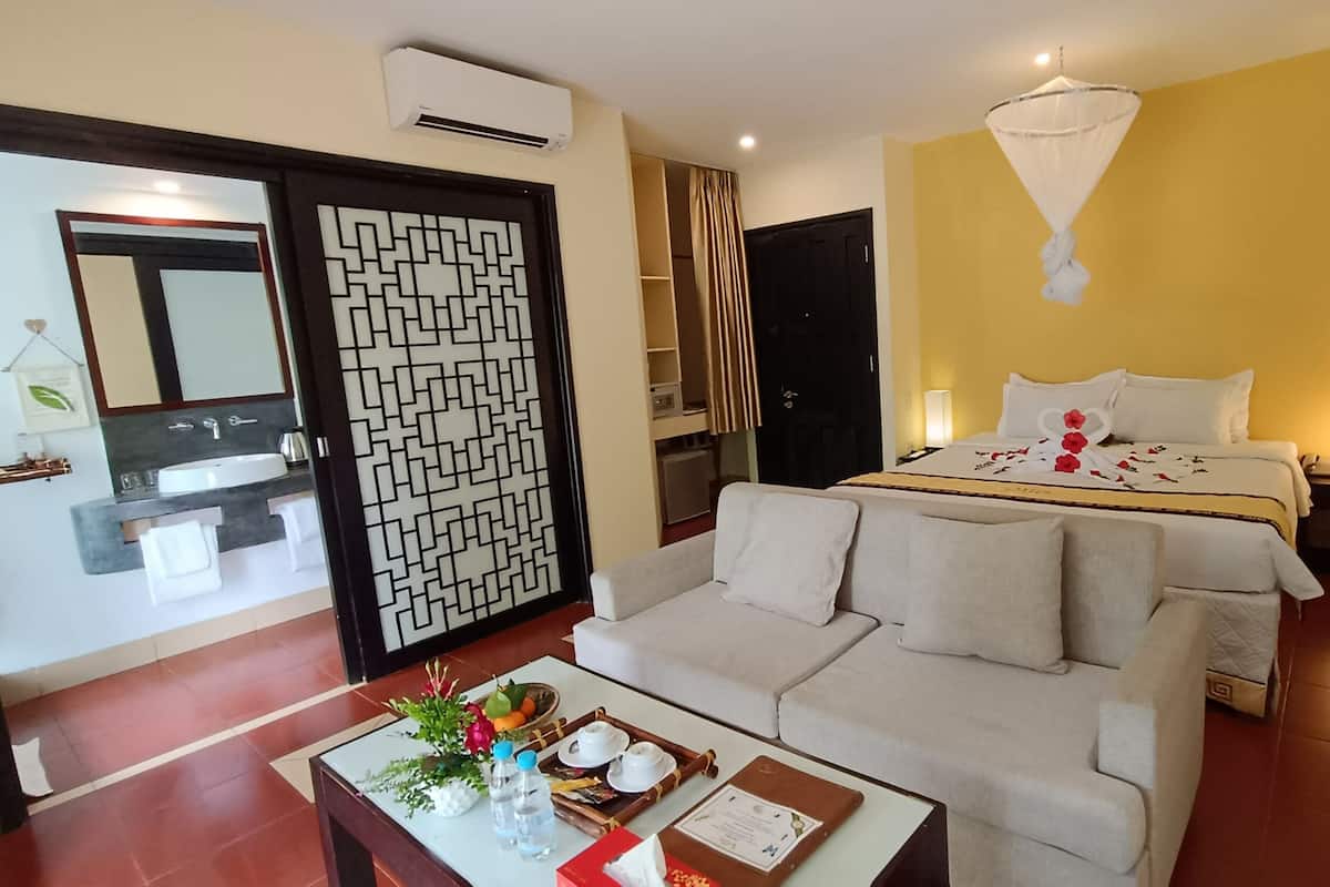 Quarto de hotel com uma cama, um sofá, uma mesa de centro com um vaso e um tabuleiro, e um banheiro com um espelho e pia.