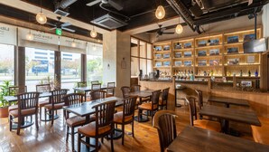 Interior de restaurante com mesas e cadeiras de madeira, área de bar com prateleiras cheias de garrafas e uma parede com quadros emoldurados.