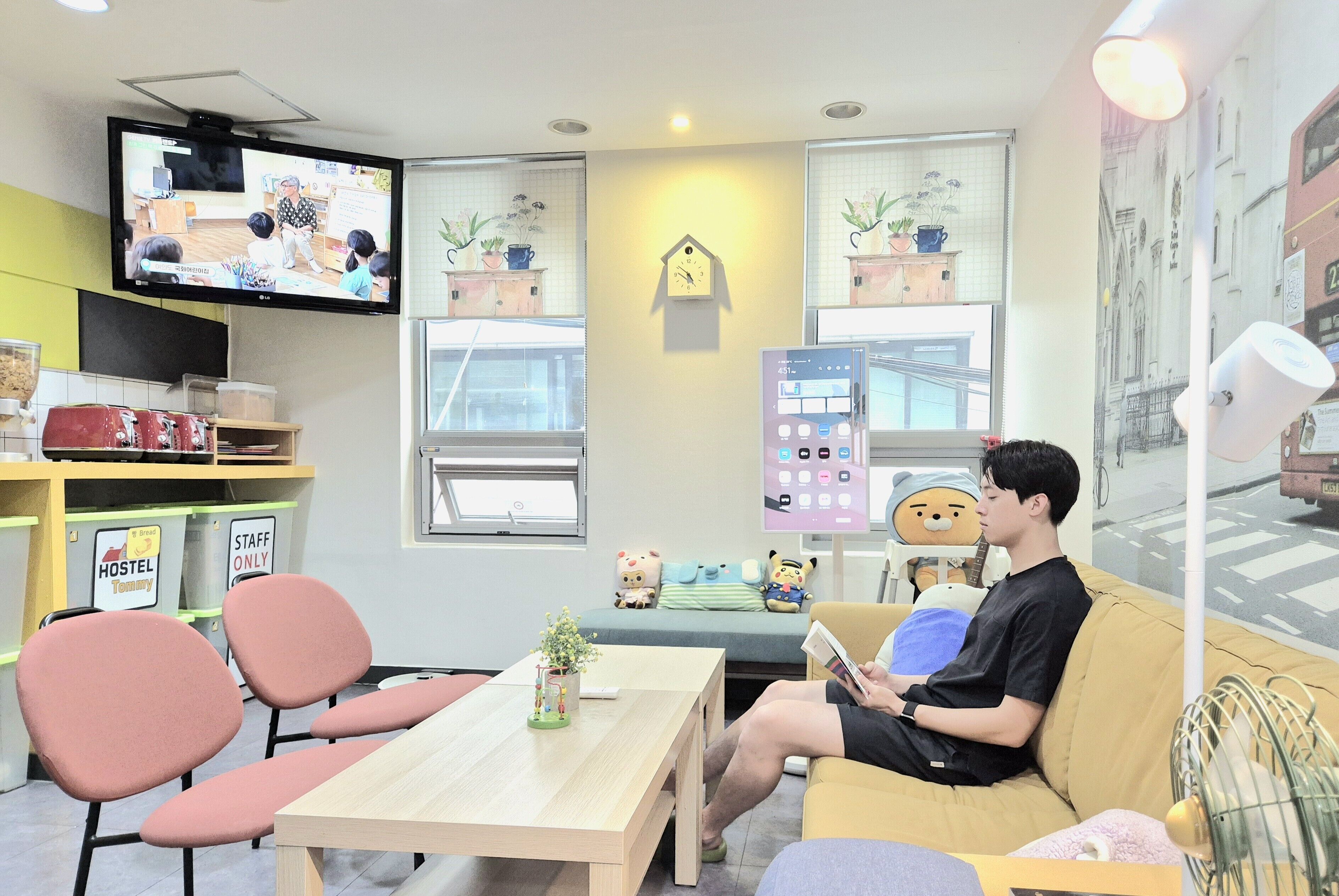 Quarto com televisão, relógio, janela com vista para um edifício, sofá e mesa com uma planta.