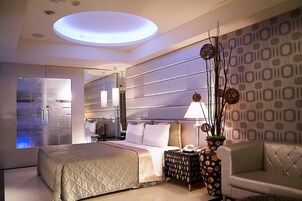 Quarto de hotel moderno com uma cama grande, mesa de cabeceira com abajur, um vaso decorativo e um sofá.