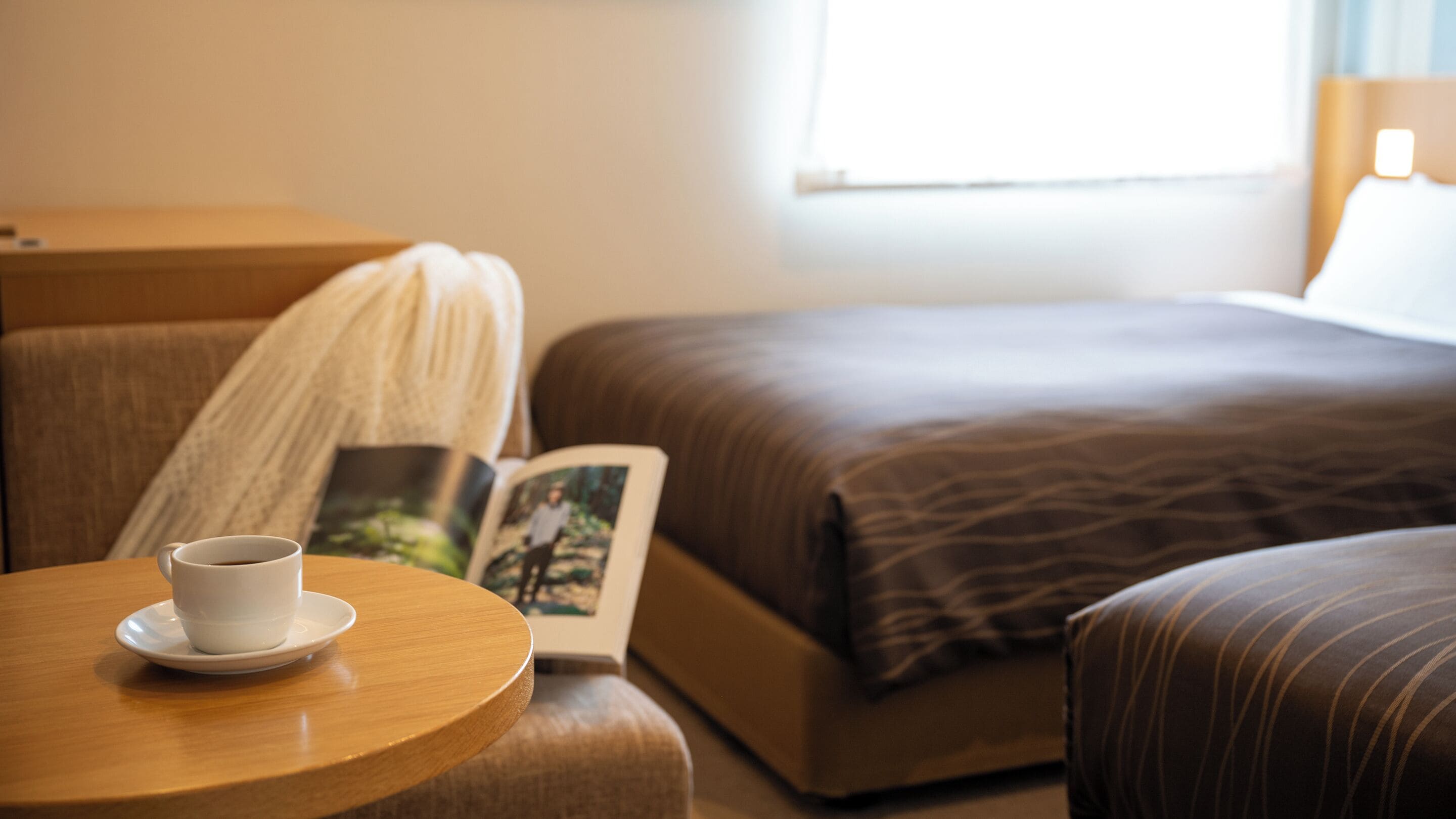 Quarto de hotel com cama, mesa de cabeceira com uma xícara de café, um livro e janela com luz natural.