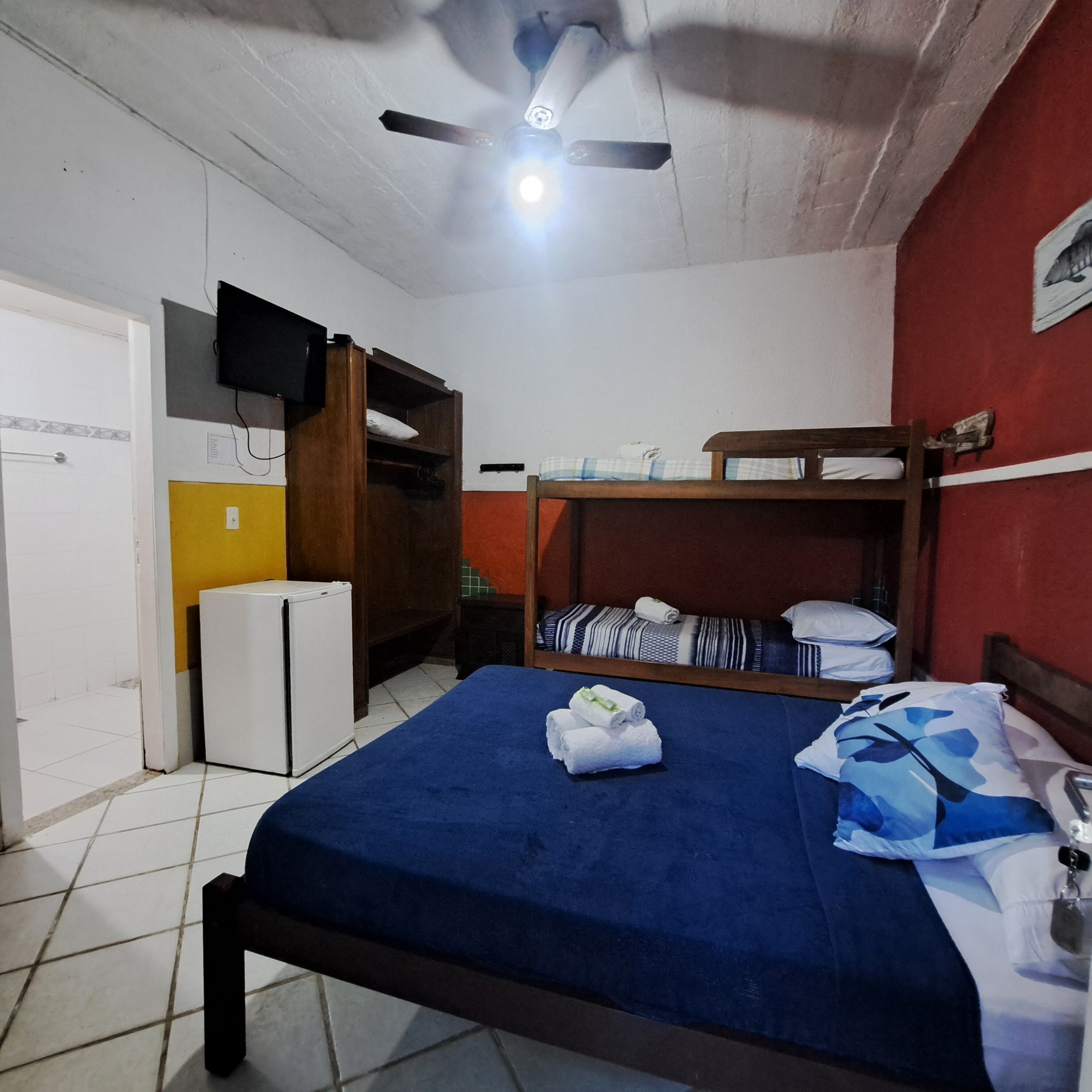 Quarto com cama, pequeno frigorífico, televisão e ventoinha no teto.