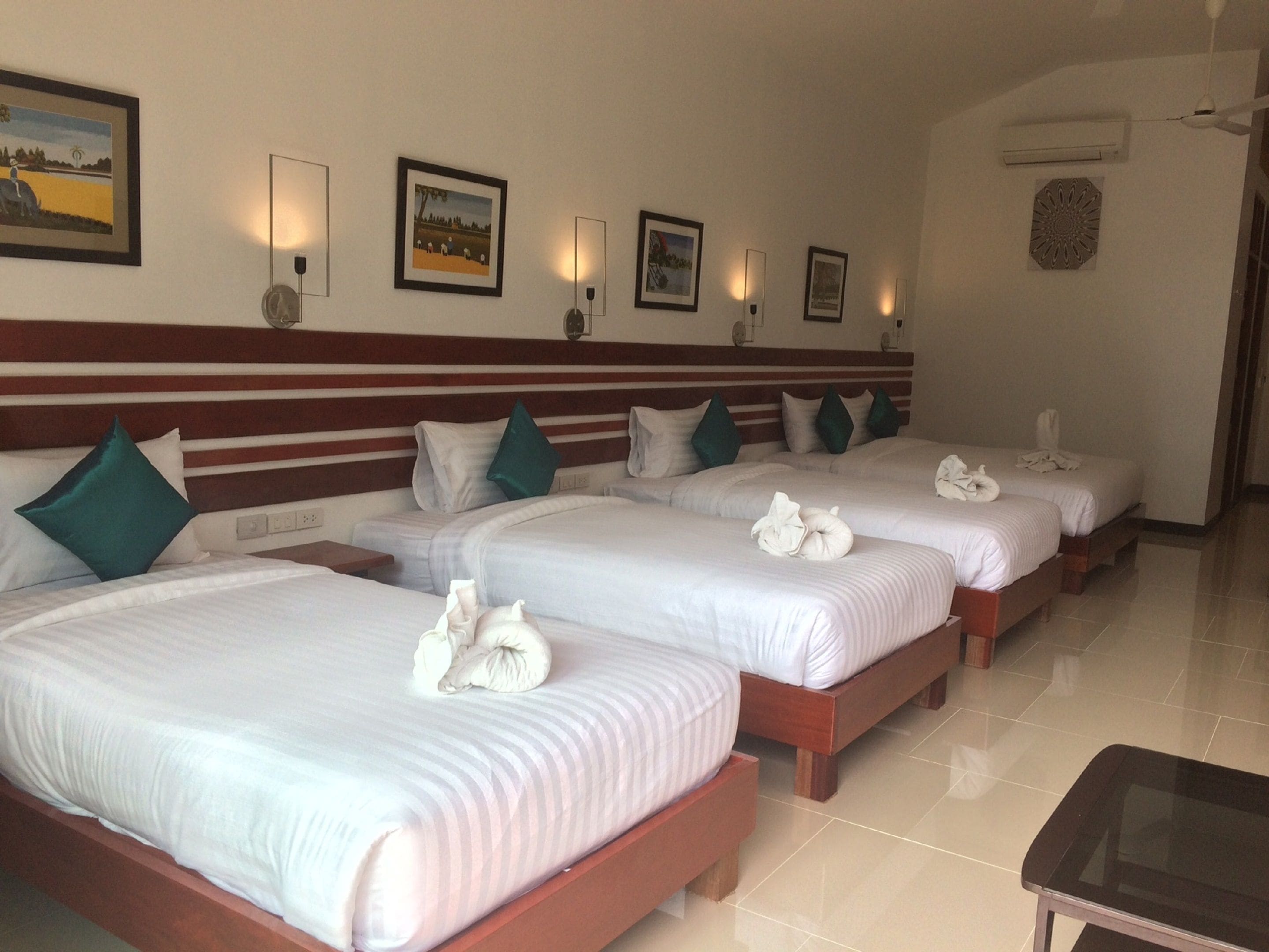 Quarto de hotel com quatro camas, cada uma com roupa de cama branca e almofadas decorativas. Há luminárias fixadas na parede e quadros acima das camas.