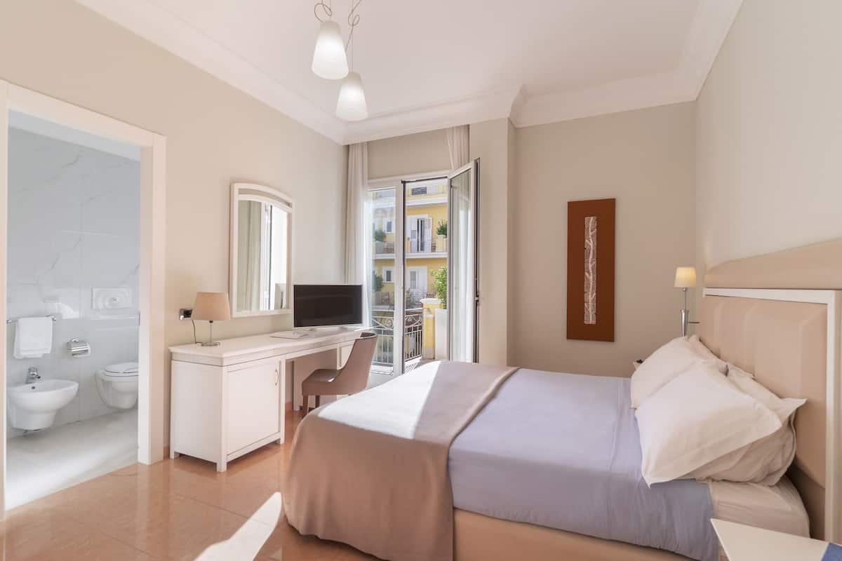 Quarto de hotel com cama, escrivaninha, televisão, banheiro com bidê e varanda com vista para prédios.
