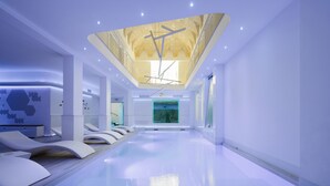 Piscina interior moderna com área de observação central, rodeada por espreguiçadeiras e teto com luminária geométrica.