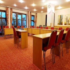 Uma sala de conferências com uma mesa comprida, cadeiras vermelhas e janelas amplas com cortinas.