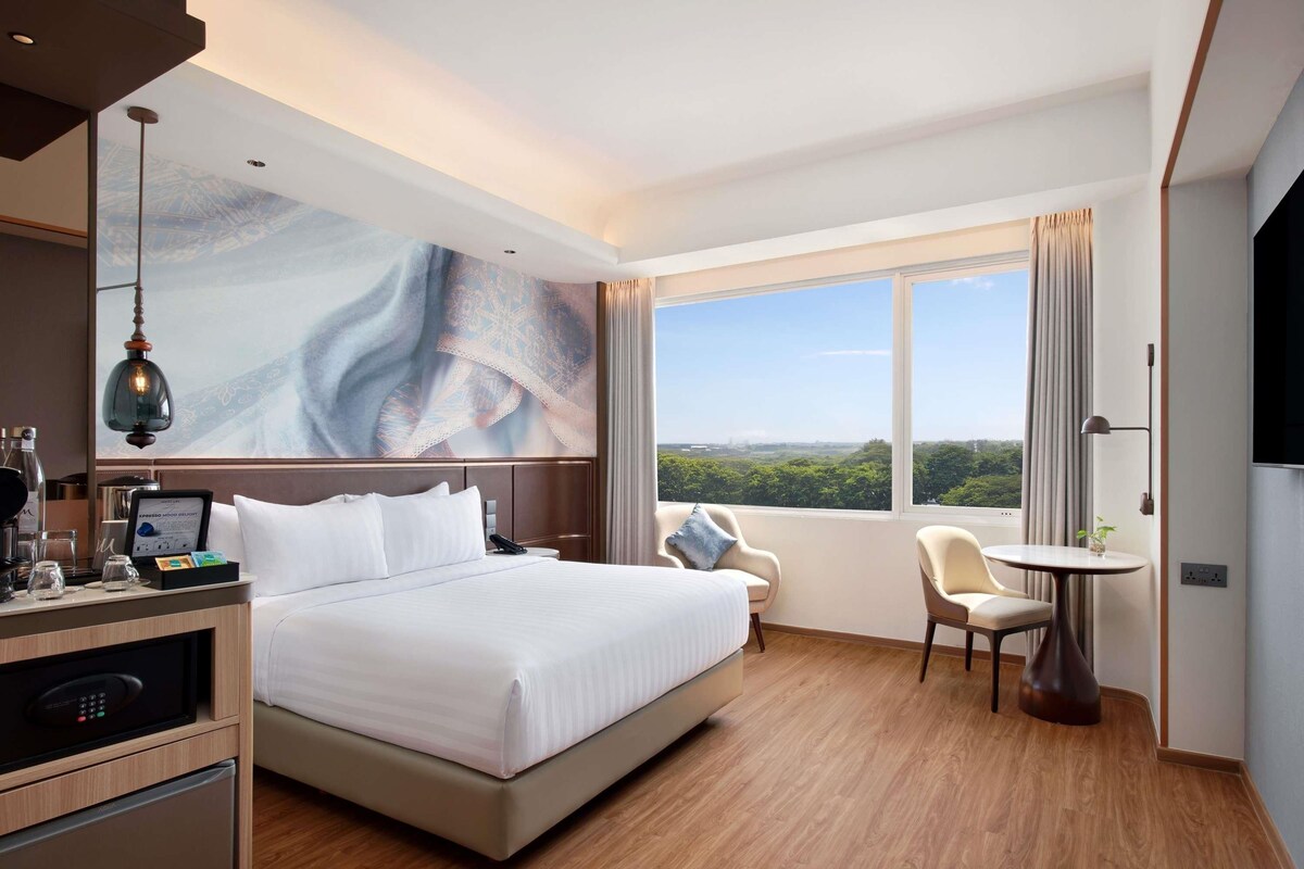 Quarto de hotel com cama, secretária com cafeteira, cadeira, mesa pequena e janela grande com vista.