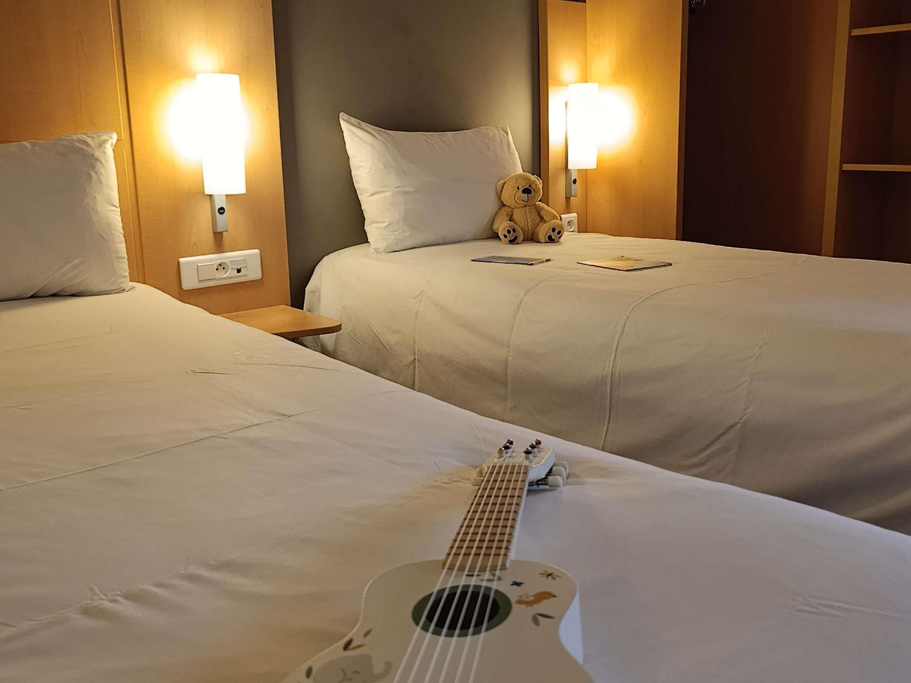 Quarto de hotel com duas camas, uma mesa de cabeceira, um abajur, um urso de pelúcia e uma guitarra.