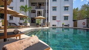 Piscina translúcida com espreguiçadeiras e guarda-sóis, um edifício branco com varandas, e uma placa indicando o Hotel 9D.