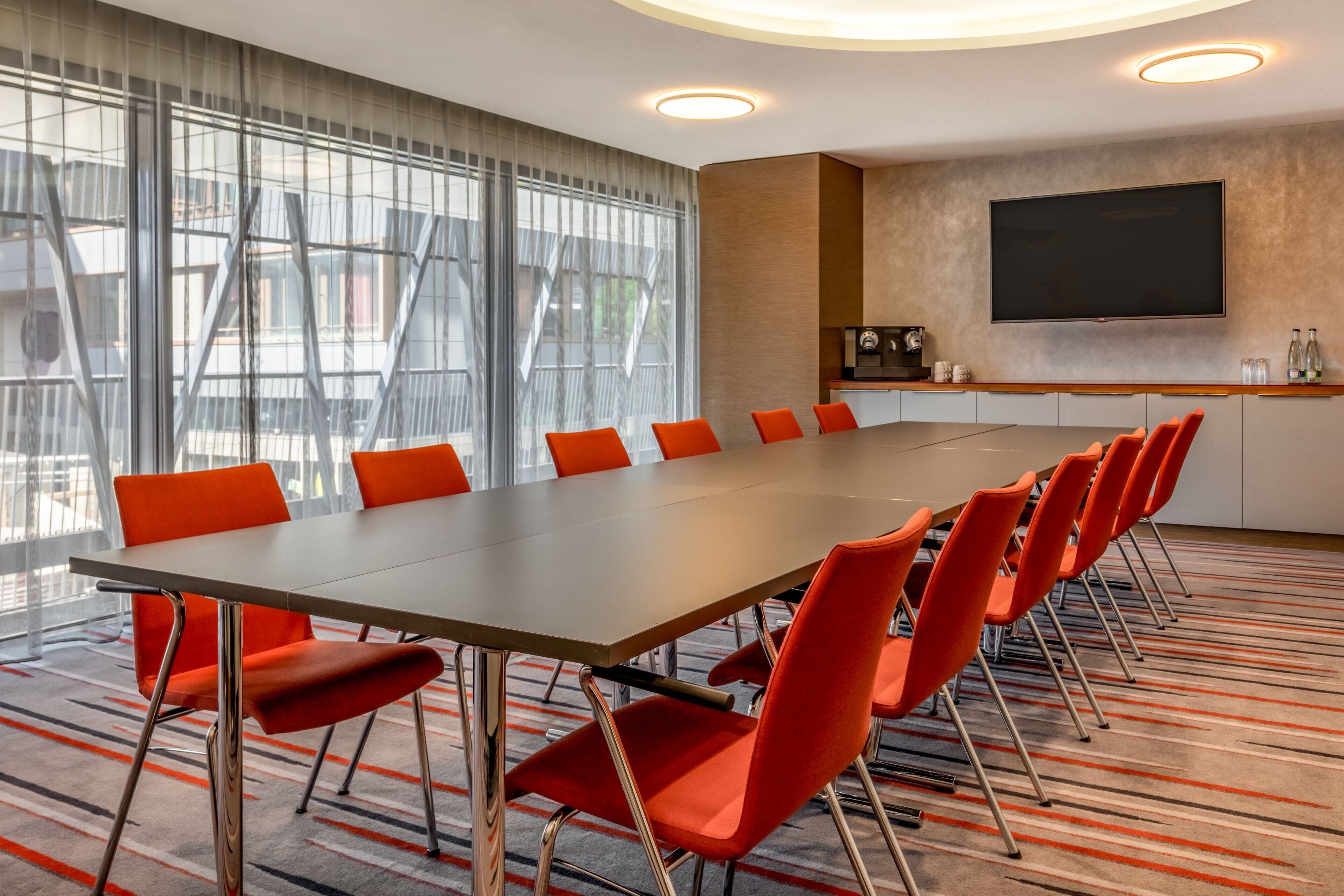 Sala de conferências com uma mesa comprida e cadeiras laranja, janelas amplas e uma televisão de tela plana na parede.