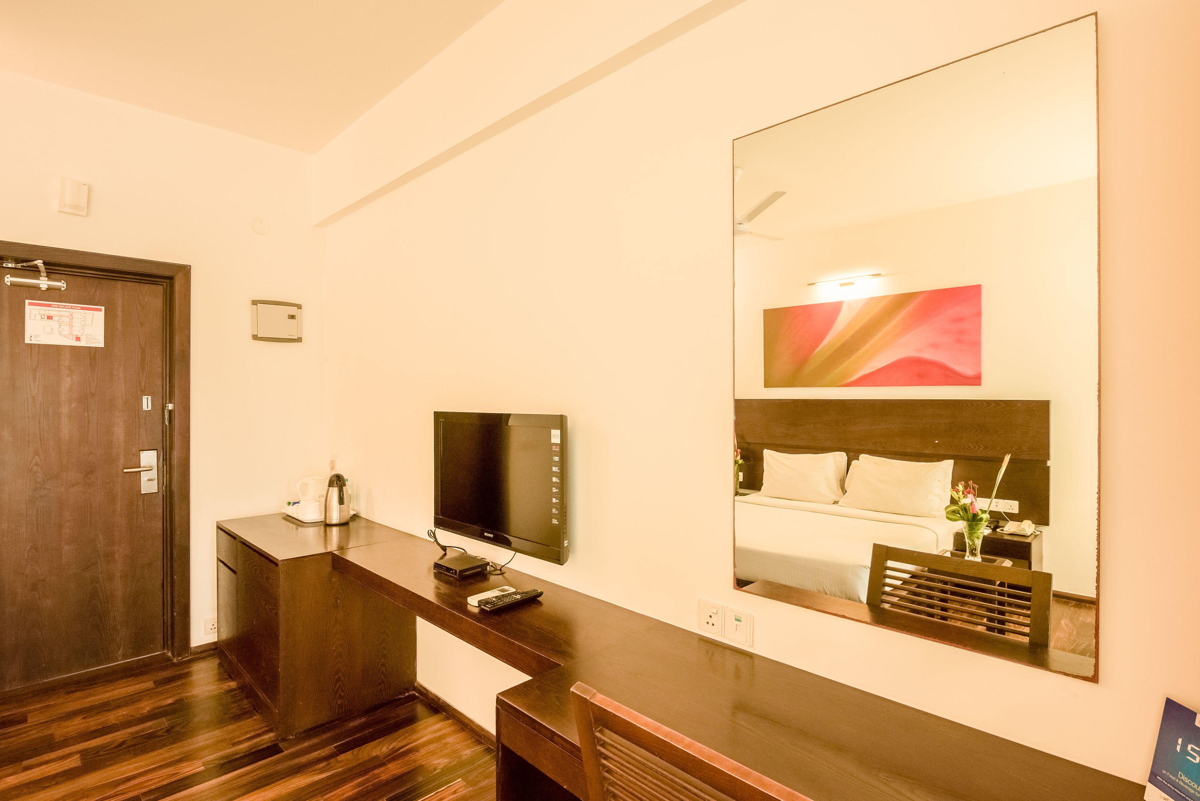 Quarto de hotel com uma escrivaninha de madeira, uma televisão de tela plana, um espelho grande e uma cama com roupa de cama branca.