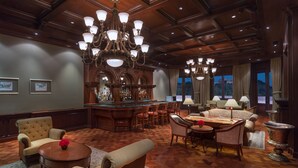 Uma área de bar com balcão de madeira, vários banquinhos e um candelabro. Há também algumas cadeiras e um sofá dispostos em torno de uma mesa de centro.