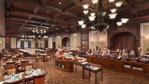 Ampla área de refeições de hotel, com várias mesas e cadeiras, um buffet e elegantes lustres.