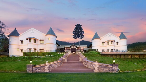 Um edifício semelhante a um castelo, com entrada central, degraus de pedra e um gramado ao redor.