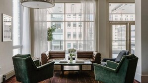 Quarto de hotel moderno com uma janela grande, área de estar com um sofá e cadeiras, e uma mesa de centro com um vaso de flores.