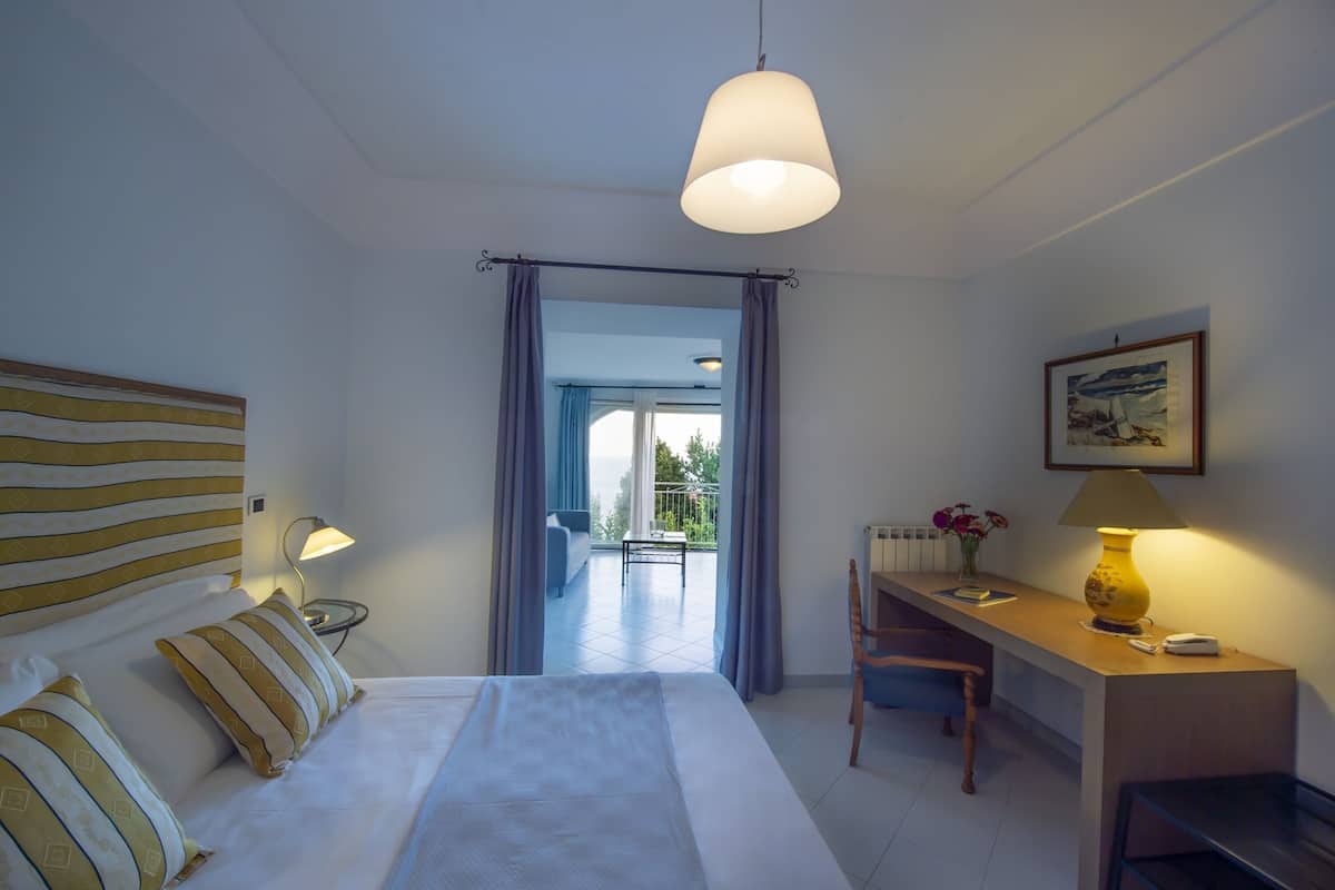 Quarto de hotel com uma cama, abajures, uma mesa de trabalho com luminária, uma cadeira e vista para uma varanda.