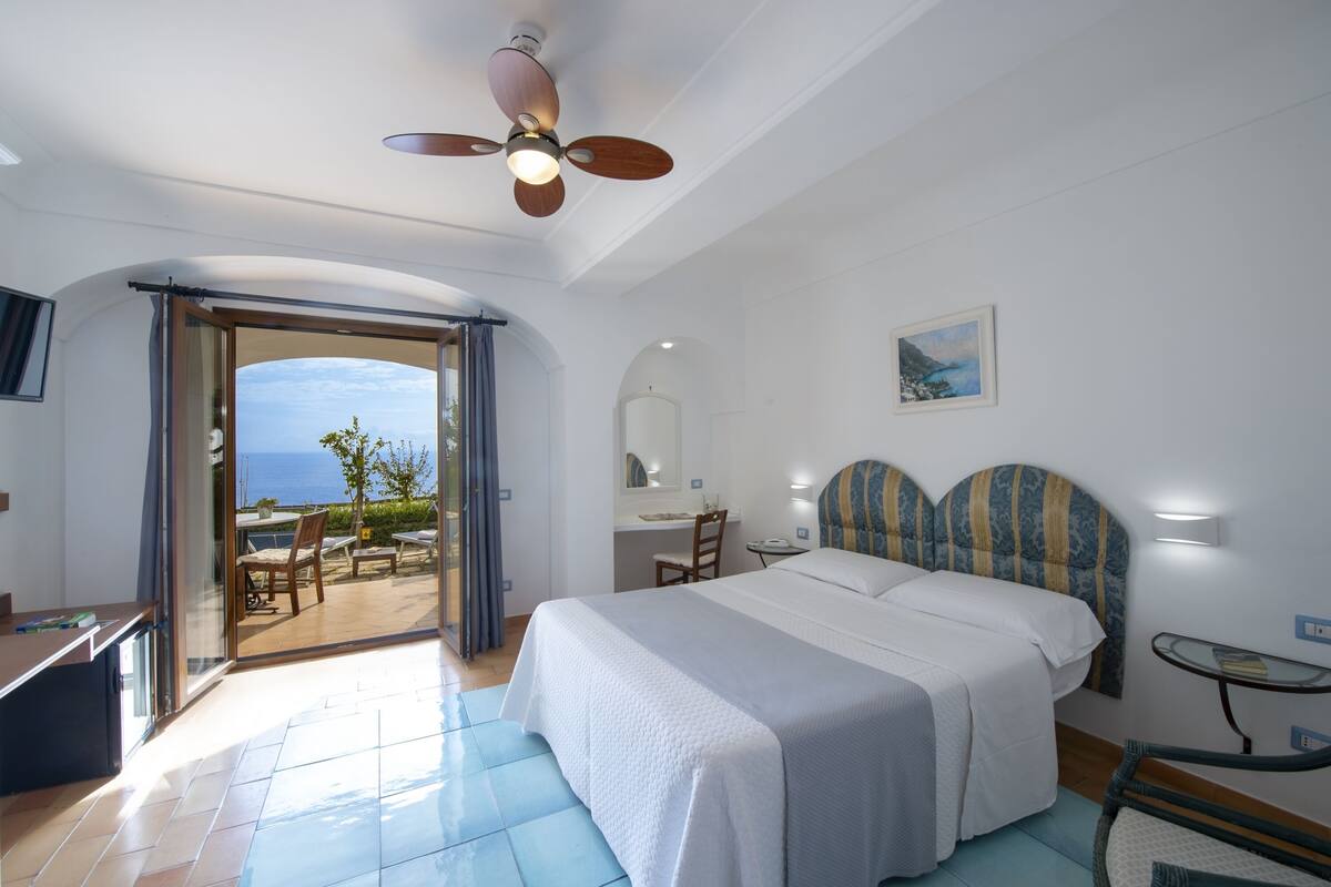 Quarto de hotel com uma cama, uma escrivaninha, uma cadeira, uma mesinha e um ventoinha de teto, além de vista para o mar.