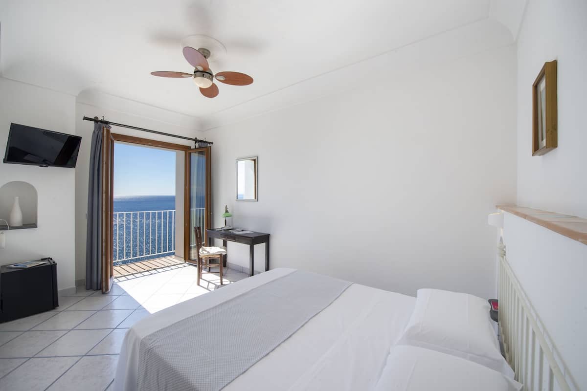 Quarto de hotel com cama, escrivaninha, cadeira, televisão e varanda com vista para o mar.