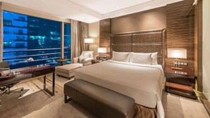 Quarto de hotel moderno com uma cama grande, uma mesa, uma cadeira e vista para a cidade à noite.