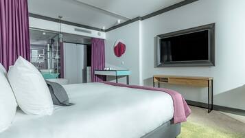 Quarto de hotel moderno com uma cama grande, uma mesa de trabalho, televisão e banheiro visível por meio de um painel de vidro.