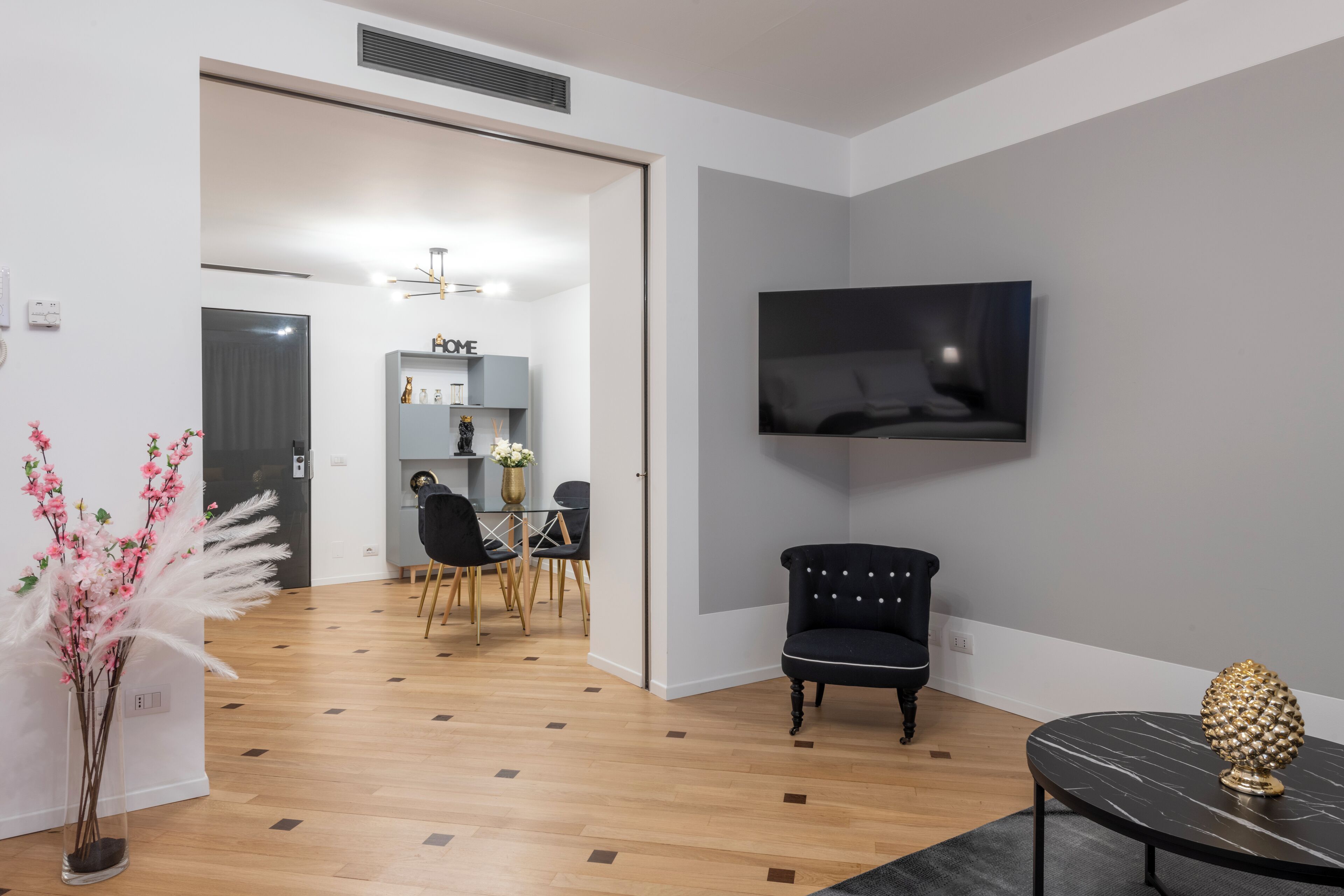 Uma sala de estar moderna com televisão de tela plana na parede, um espaço de jantar com mesa e cadeiras, e um vaso decorativo com flores cor-de-rosa.