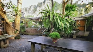 Uma área de estar ao ar livre rústica, com mesa e banco de madeira, cercada por folhagem exuberante e uma cerca de bambu.