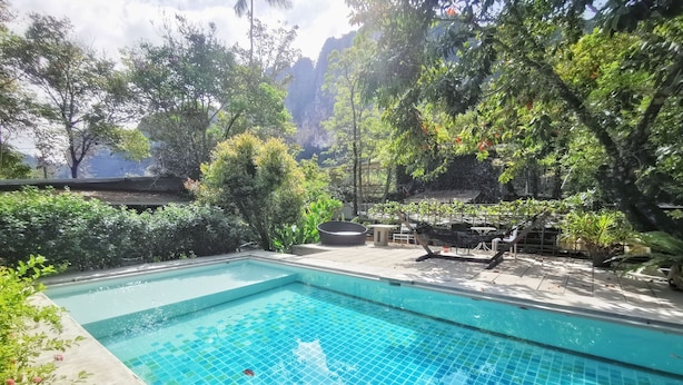 Uma piscina retangular com água azul cristalina, rodeada por vegetação exuberante e vista para um penhasco rochoso.