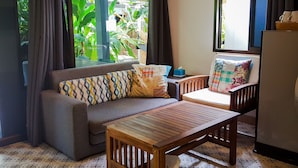 Um quarto aconchegante com sofá, mesa de madeira e uma cadeira. Há uma janela com cortinas, uma mesinha com caixa de lenços e uma geladeira.