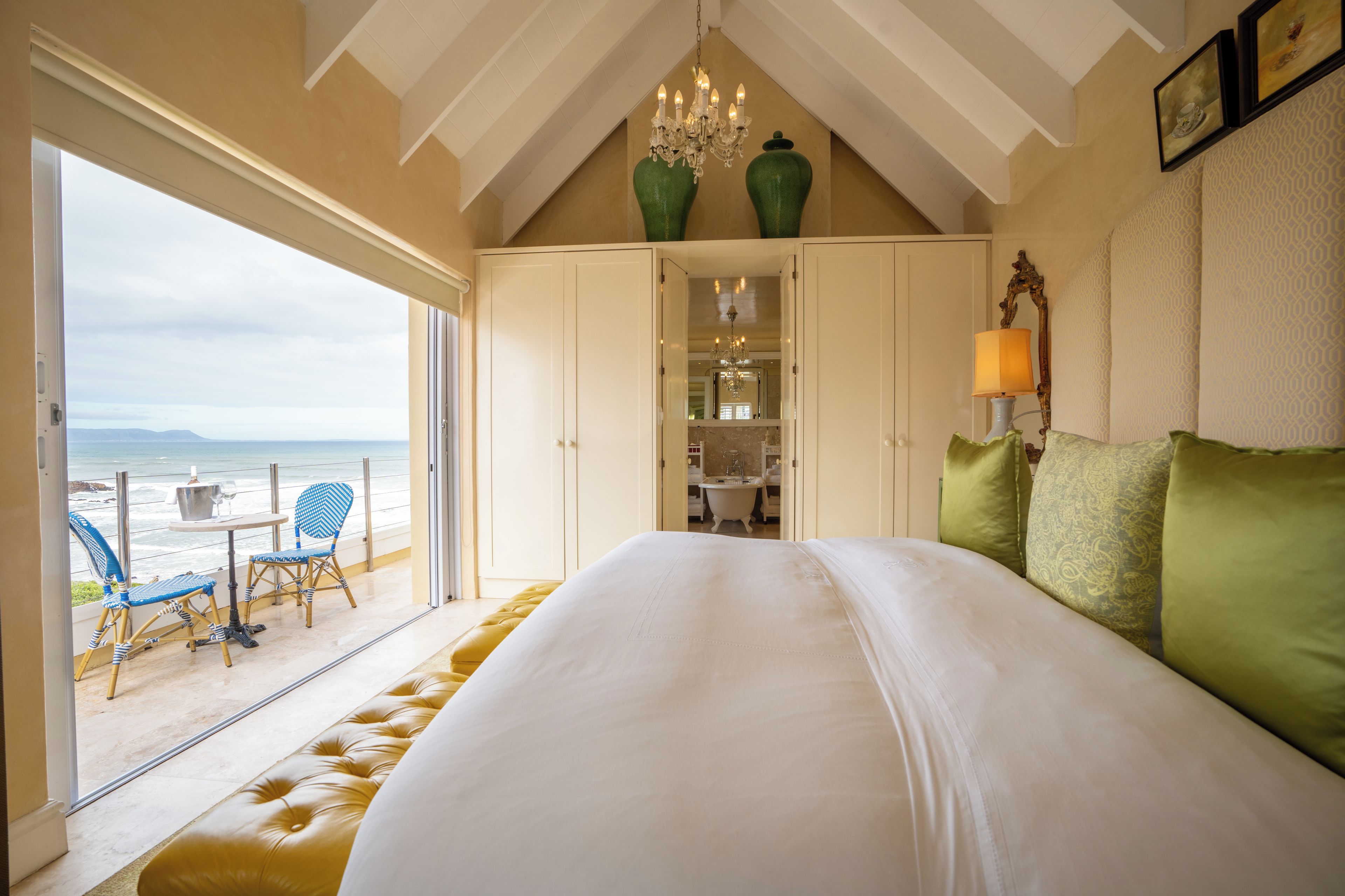 Quarto com uma cama grande, uma varanda com mesa e cadeiras, e vista para o mar.