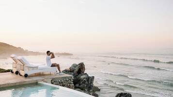 Homem fotografando com uma câmera, sentado em uma espreguiçadeira à beira da piscina com vista para o oceano.
